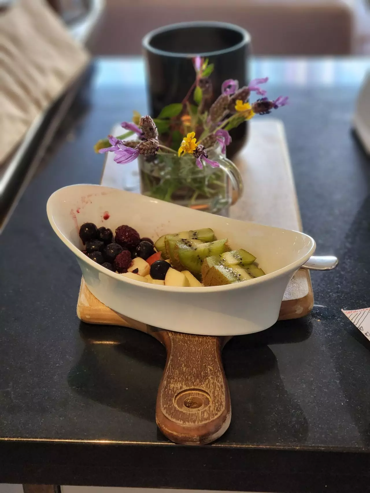 Unha cunca de froitas en rodajas sobre unha táboa de madeira diante dun vaso e flores e unha vela.